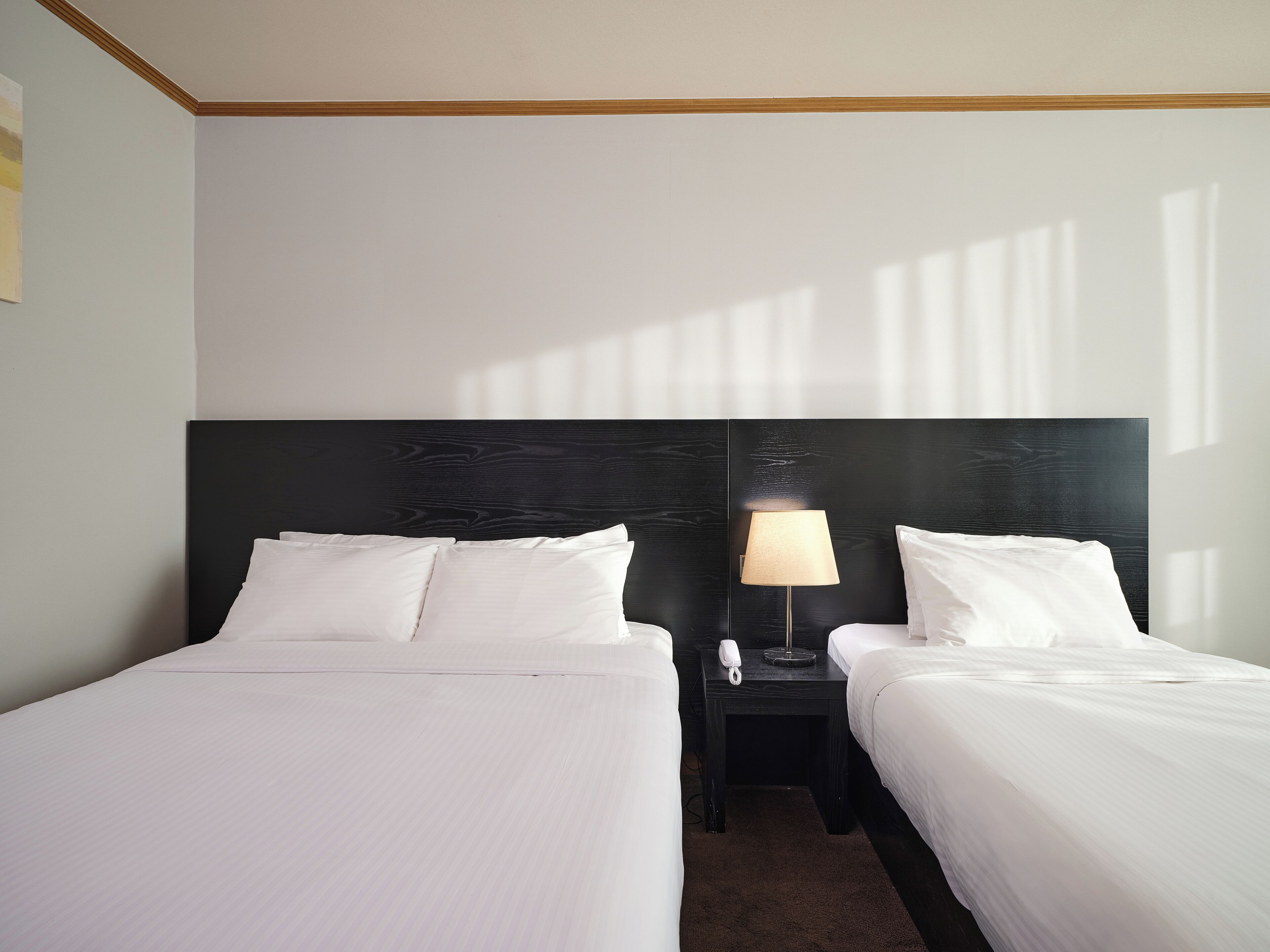 Quarto de hotel com duas camas, uma mesa de cabeceira com abajur e uma parede lisa.