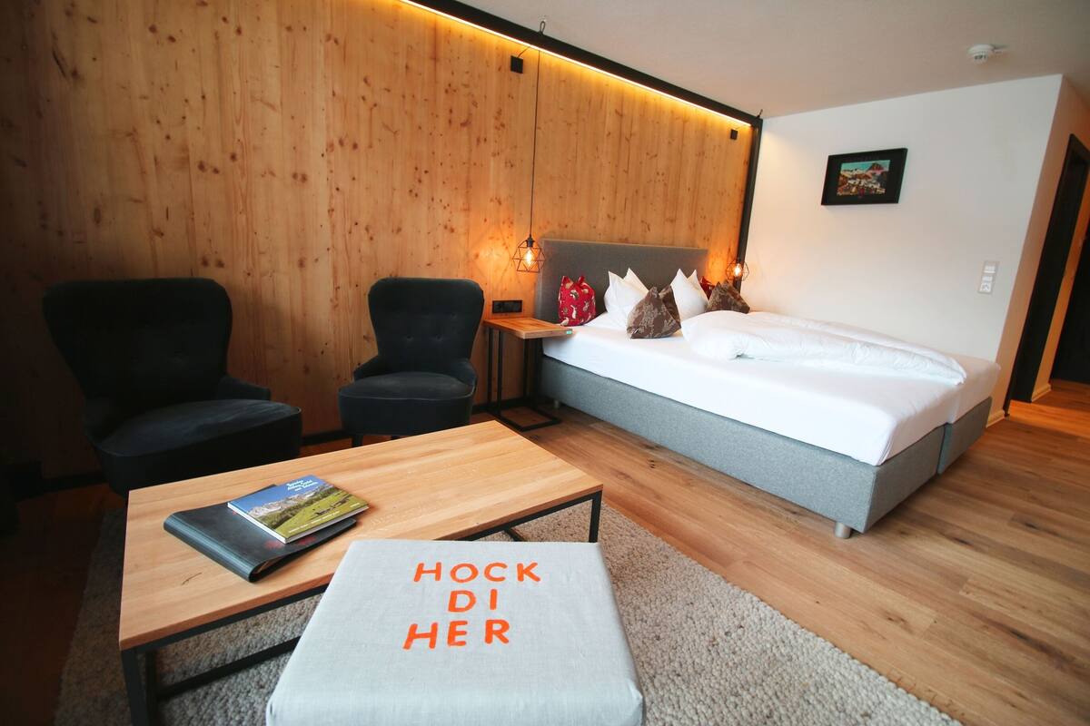 Quarto de hotel moderno com parede de madeira, cama com roupa de cama branca, duas cadeiras pretas, mesa de madeira com um livro e um tapete pequeno com a inscrição "Hock di Her".