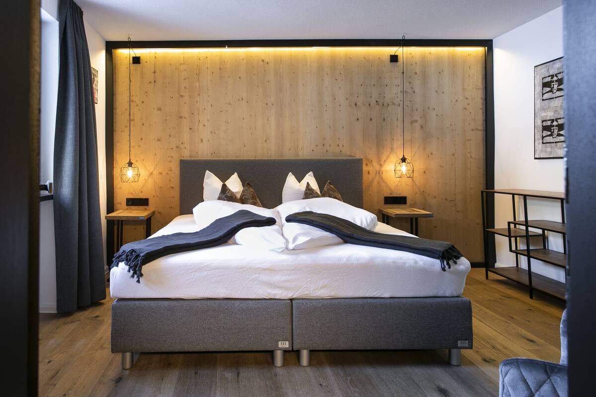 Quarto moderno com cabeceira de madeira, cama grande com roupa de cama branca, duas luminárias de mesa e piso de madeira.