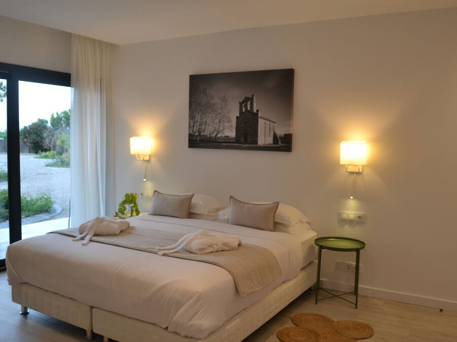 Quarto de hotel com uma cama grande, duas luminárias de cabeceira, um quadro na parede e vista pela porta de vidro.