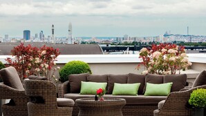 Um terraço no telhado com móveis de vime, um sofá marrom com almofadas verdes e vista para o horizonte da cidade.