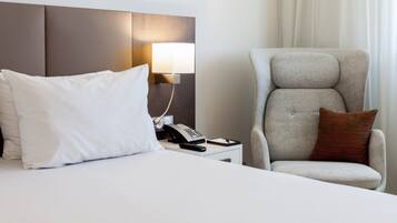 Quarto de hotel com uma cama, mesa de cabeceira com luminária, telefone e uma cadeira com almofada.