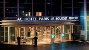Exterior do AC Hotel Paris Le Bourget Airport, com fachada moderna em vidro, sinalização iluminada e uma entrada convidativa.