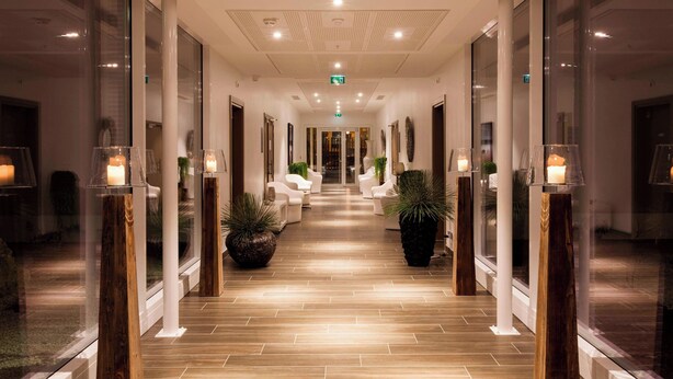 Um corredor de hotel com piso de madeira, cadeiras brancas, plantas em vasos e pilares de vidro iluminados com velas.
