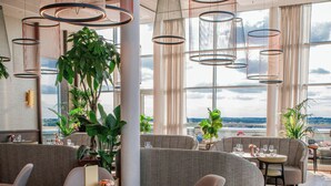 Um restaurante moderno com pé-direito alto, luminárias pendentes e áreas de estar com vista para a cidade.