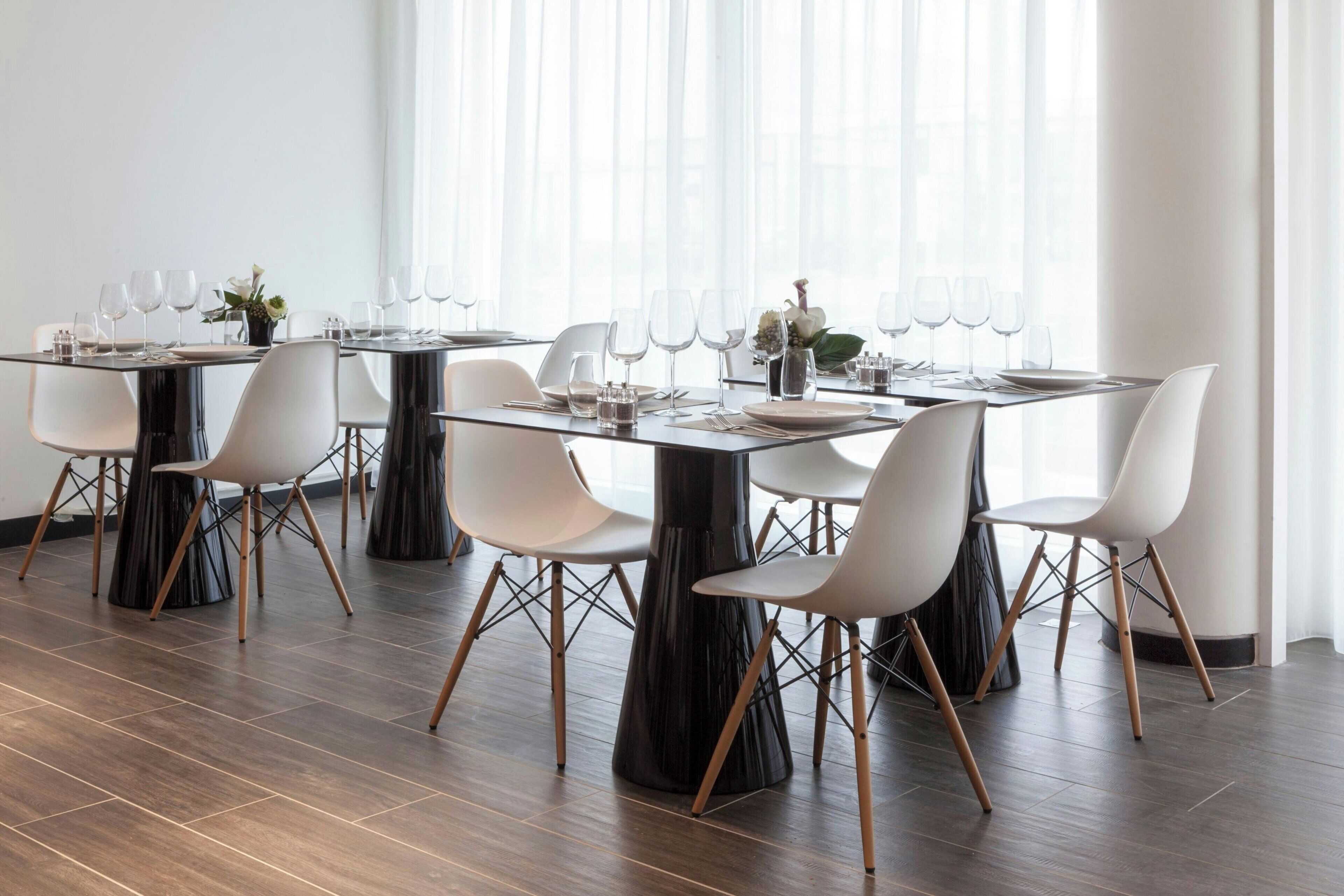 Uma sala de jantar moderna com mesas preparadas para quatro pessoas, cada uma com cadeiras brancas e um vaso central.