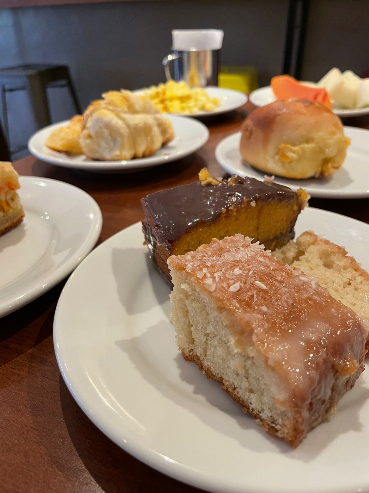 Uma mesa com pratos de doces, incluindo uma fatia de bolo com cobertura brilhante e um pedaço de pão com cobertura açucarada.