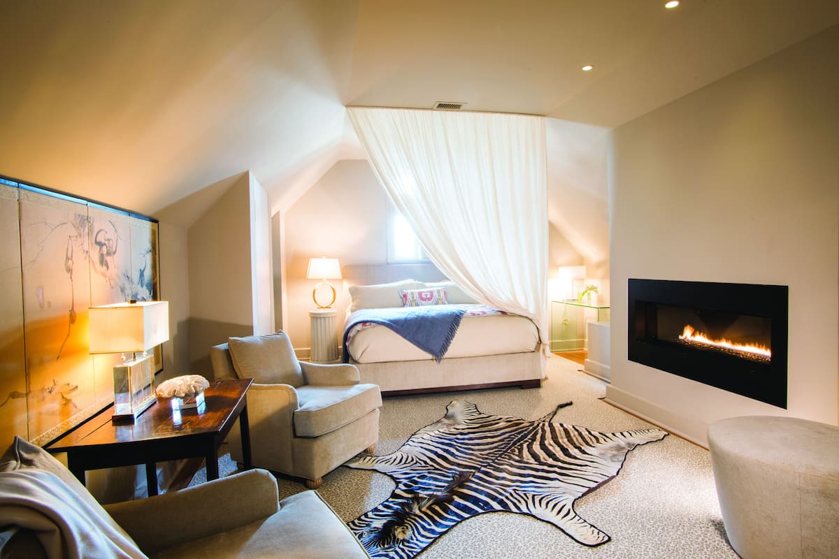 Quarto de hotel moderno com uma cama, área de estar com uma poltrona e uma mesa, lareira e tapete de zebra.