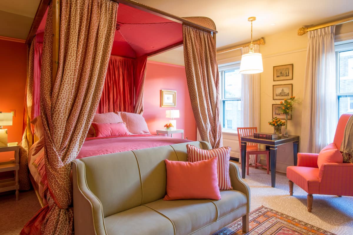 Uma cama com dossel e cortinas, roupa de cama rosa, um sofá verde com almofadas cor-de-rosa, uma cadeira de madeira, uma mesa pequena com um vaso de flores e uma janela com cortinas.