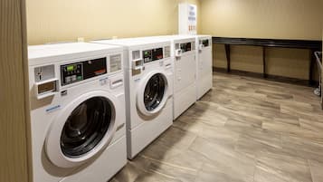 Fileira de máquinas de lavar e secar de carregamento frontal brancas em uma lavanderia de hotel, com piso ladrilhado e sinalização indicando os tipos de detergentes disponíveis.