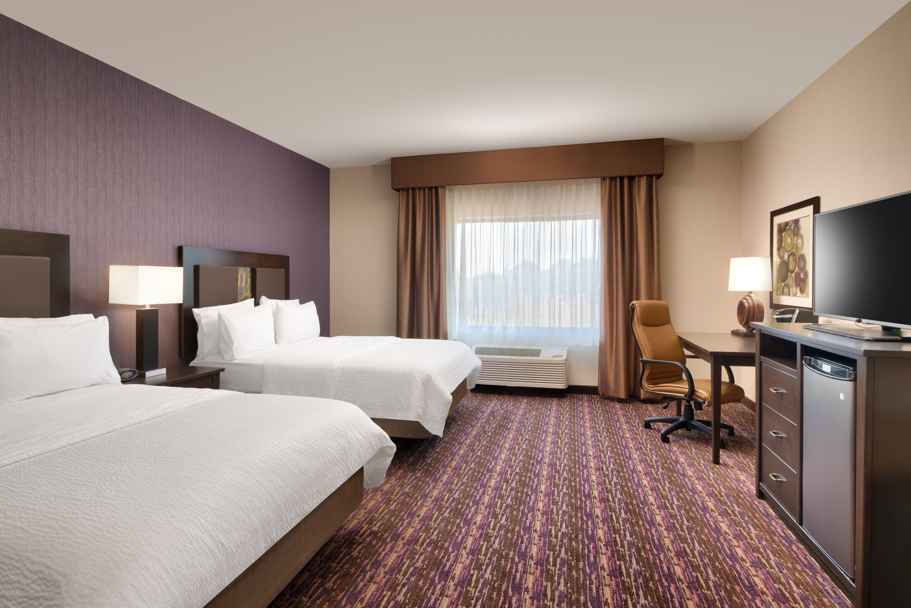 Quarto de hotel com duas camas, uma escrivaninha, uma cadeira, uma televisão e uma janela com cortinas.