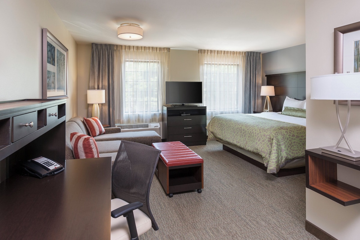 Quarto de hotel com cama, escrivaninha com telefone, cadeira, televisão e mesa de cabeceira com abajur.