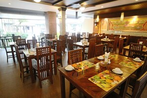 Um restaurante com mesas e cadeiras de madeira, dispostas numa ampla sala de jantar com grandes janelas que permitem a entrada de luz natural.