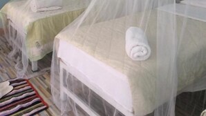 Quarto com duas camas, cada uma coberta por um mosquiteiro branco. Há uma janela com persianas e um tapete listrado visível no chão.