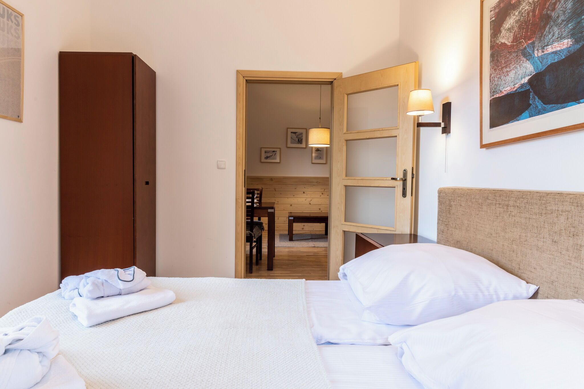 Quarto de hotel com cama, guarda-roupa, porta de madeira para uma sala de jantar e uma luminária fixada na parede.