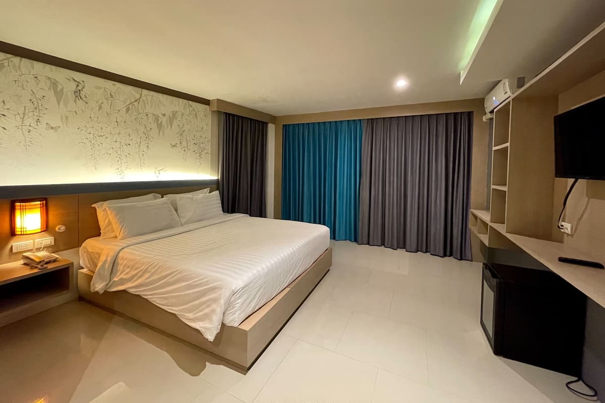 Quarto de hotel moderno com uma cama grande, mesa de cabeceira com abajur, televisão fixada na parede e uma janela ampla com cortinas.