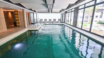 Piscina interior moderna com água cristalina, deck de madeira e vista para prédios externos através de grandes janelas.
