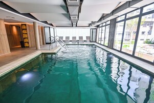 Piscina interior moderna com água cristalina, deck de madeira e vista para prédios externos através de grandes janelas.