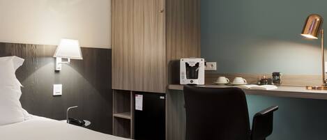 Quarto de hotel com cama, escrivaninha, cafeteira, luminária e cadeira.