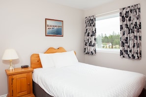 Quarto com cabeceira de madeira, colcha branca, mesa de cabeceira com abajur, quadro na parede e janela com cortinas florais.
