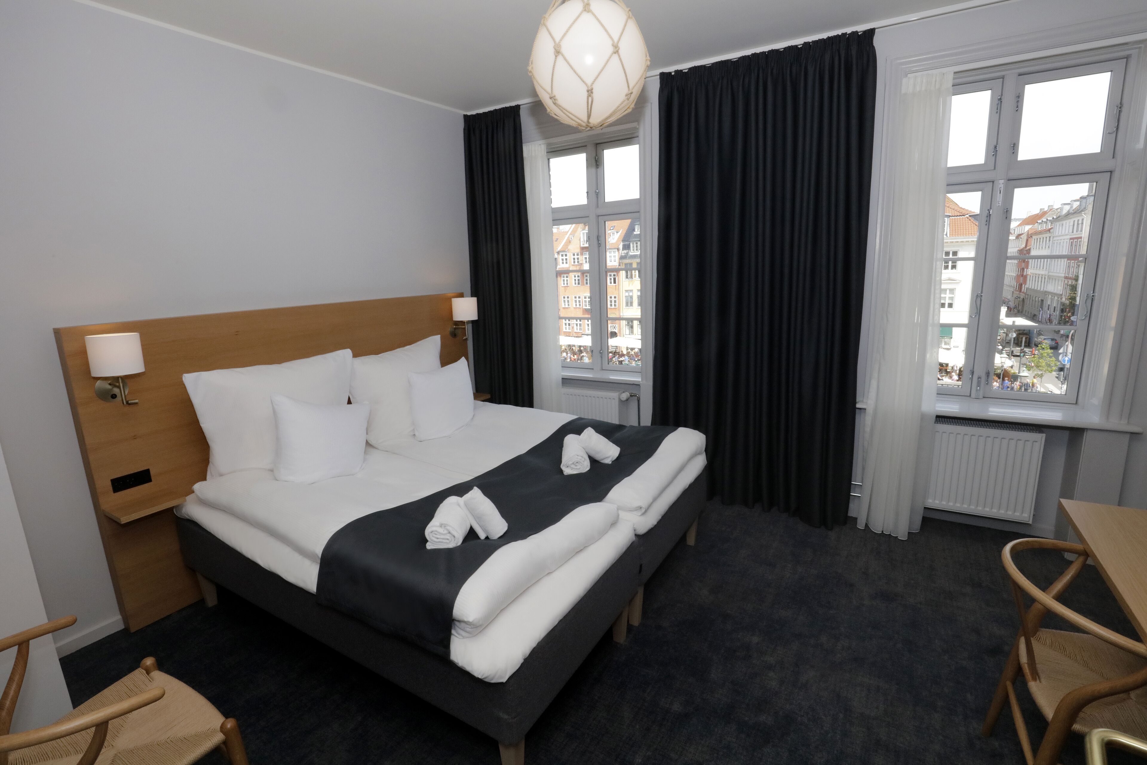 Quarto de hotel com uma cama grande, mesa de cabeceira de madeira, escrivaninha e janela com cortinas.