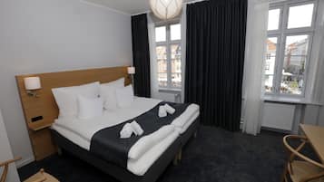 Quarto de hotel com uma cama grande, mesa de cabeceira de madeira, escrivaninha e janela com cortinas.