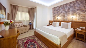 Quarto de hotel com cama grande, cabeceira de madeira, parede de tijolos decorativa, pequena mesa com um vaso de flores e sofá com manta estampada.