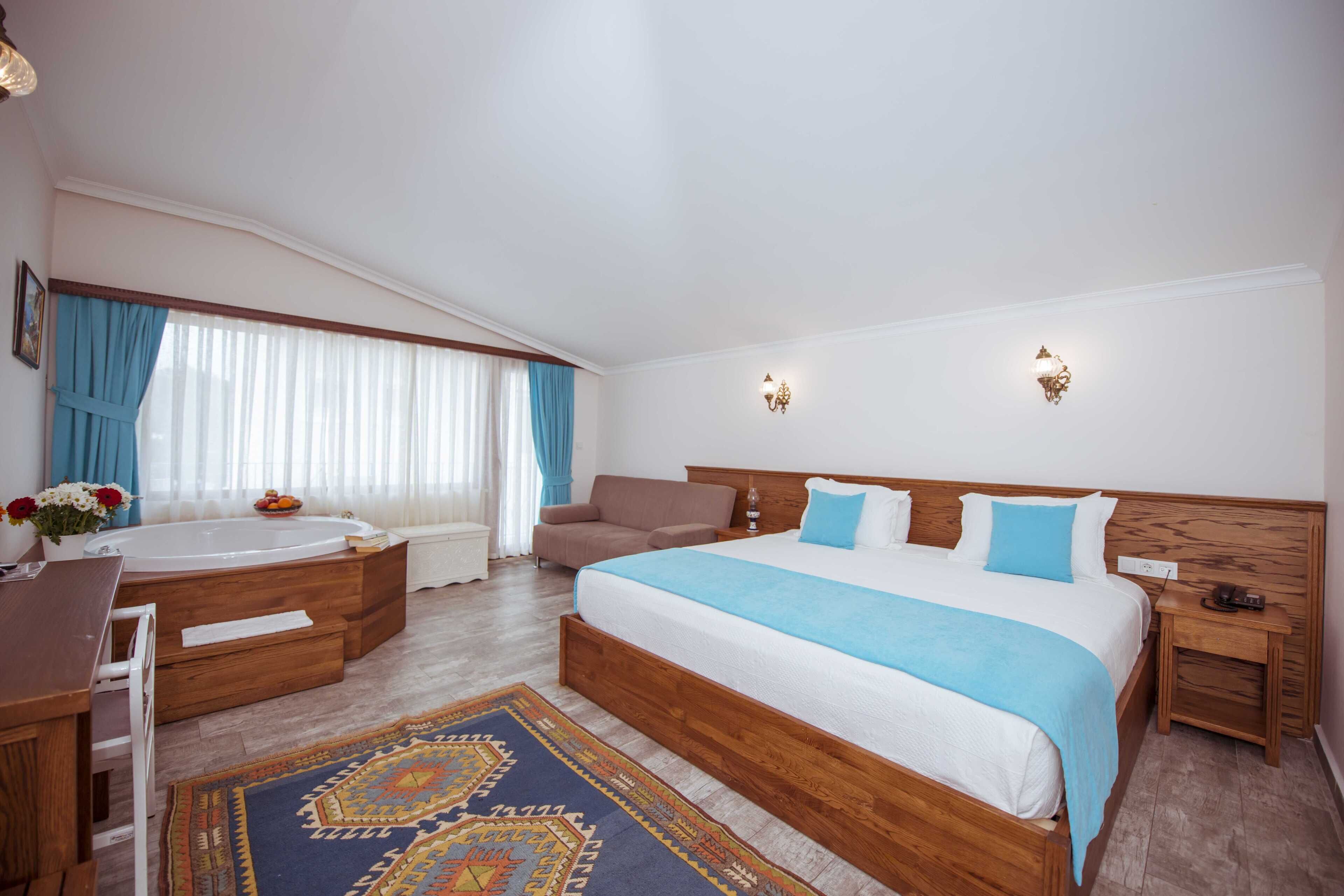 Um quarto espaçoso com uma cama grande, cabeceira de madeira, área de estar com uma poltrona, uma mesinha, uma banheira e uma janela com cortinas.
