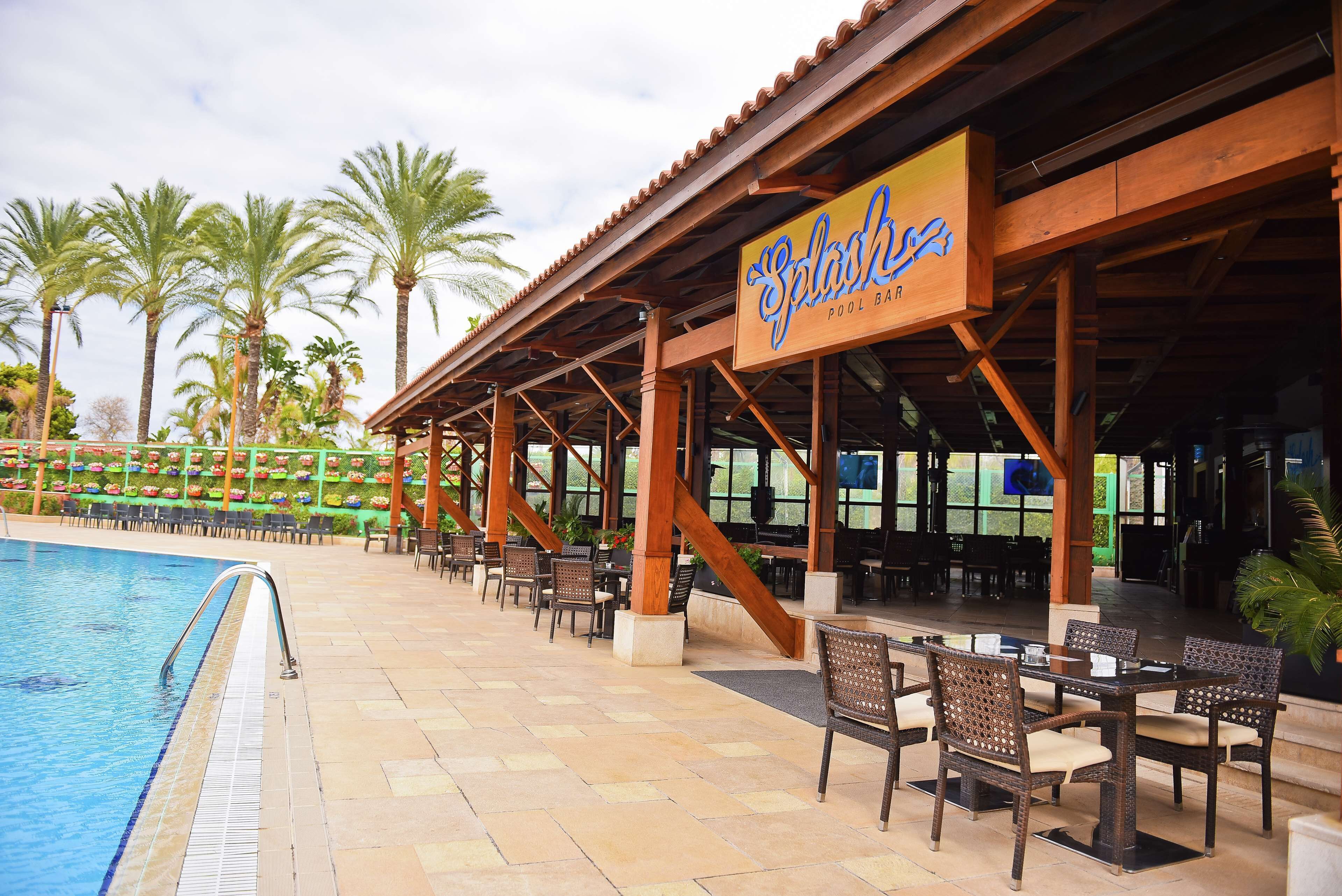 Uma área de piscina com um letreiro que diz "Splash Pool Bar", cercada por pérgulas de madeira e assentos externos.