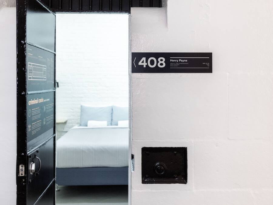 Quarto de hotel com uma cama, uma placa na parede indicando o número 408, e um pequeno painel preto na parede.