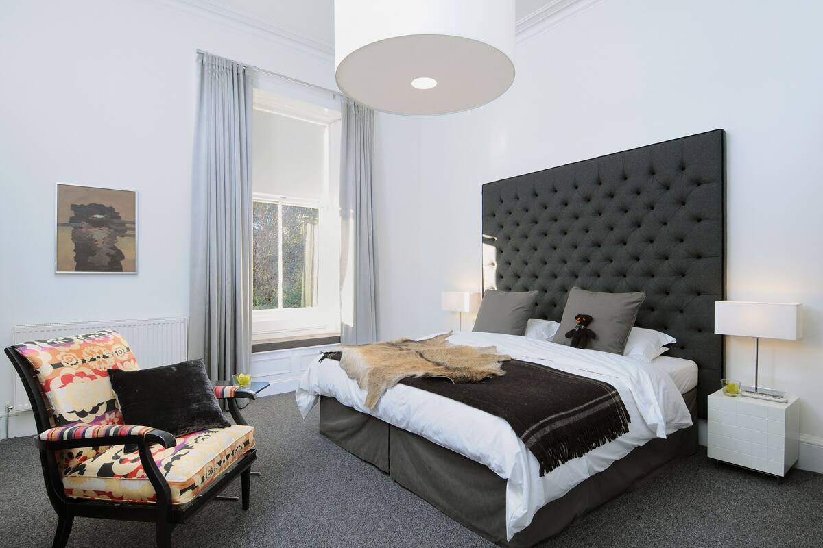 Um quarto moderno com uma cama grande, uma cabeceira estofada, uma cadeira com estampa floral e uma janela com cortinas.