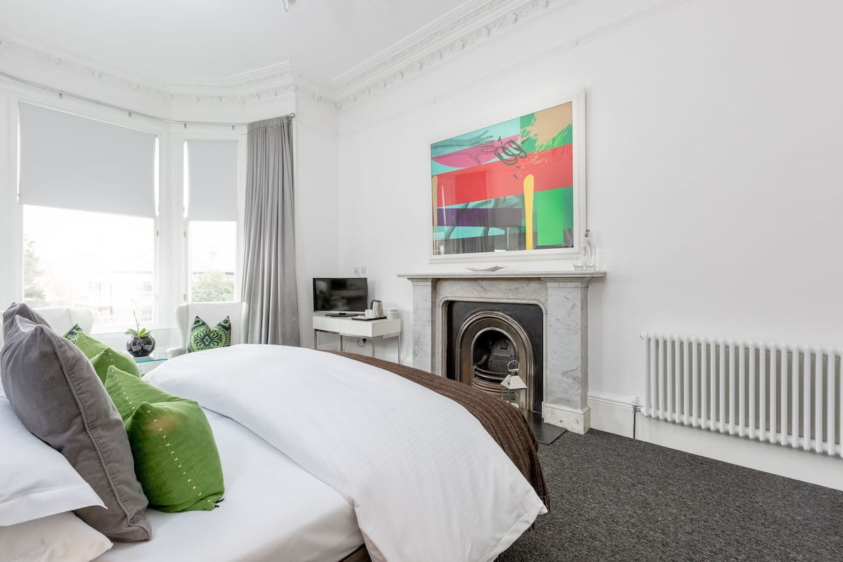 Quarto de hotel moderno com uma cama grande, lareira, escrivaninha e um quadro colorido na parede.