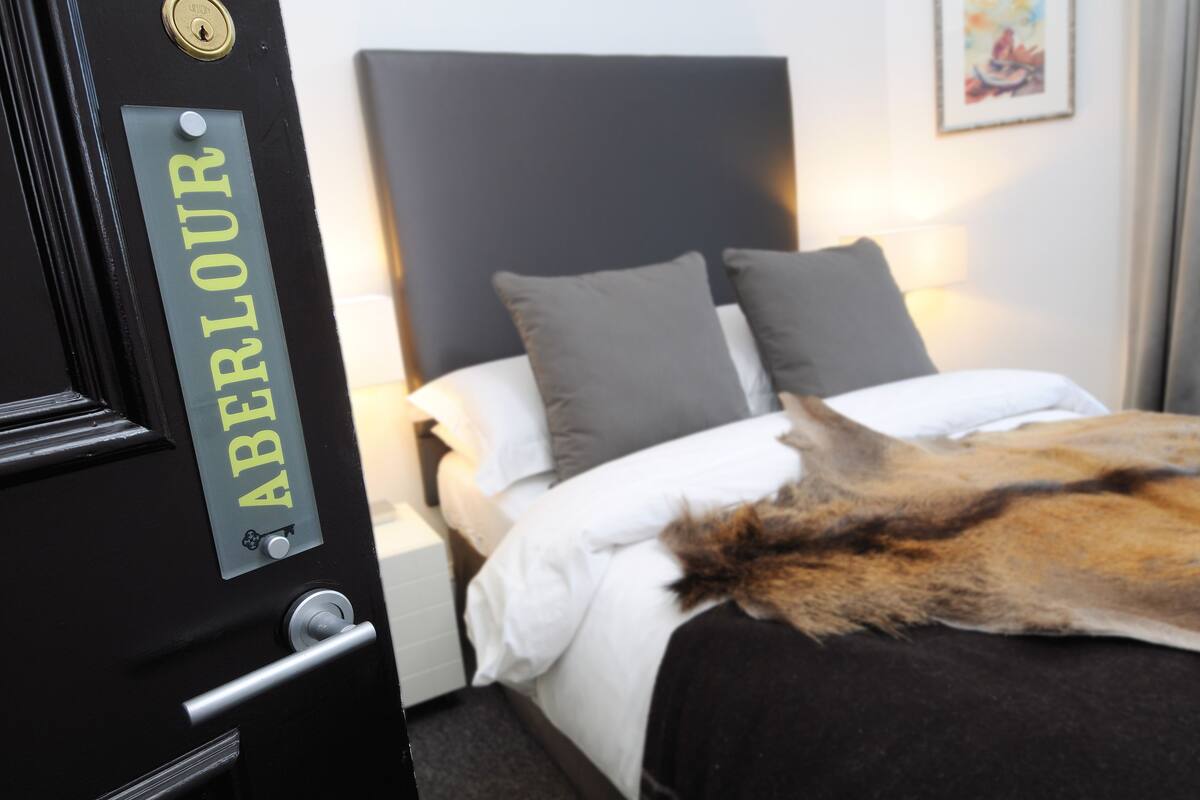 Quarto de hotel com cama, cabeceira preta e um aviso na porta que diz "ABERLOUR".