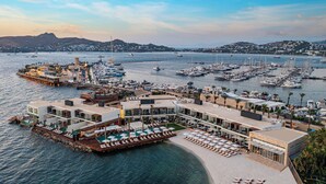 Uma marina com vários barcos, um restaurante à beira-mar com área externa e vista para as colinas ao redor.