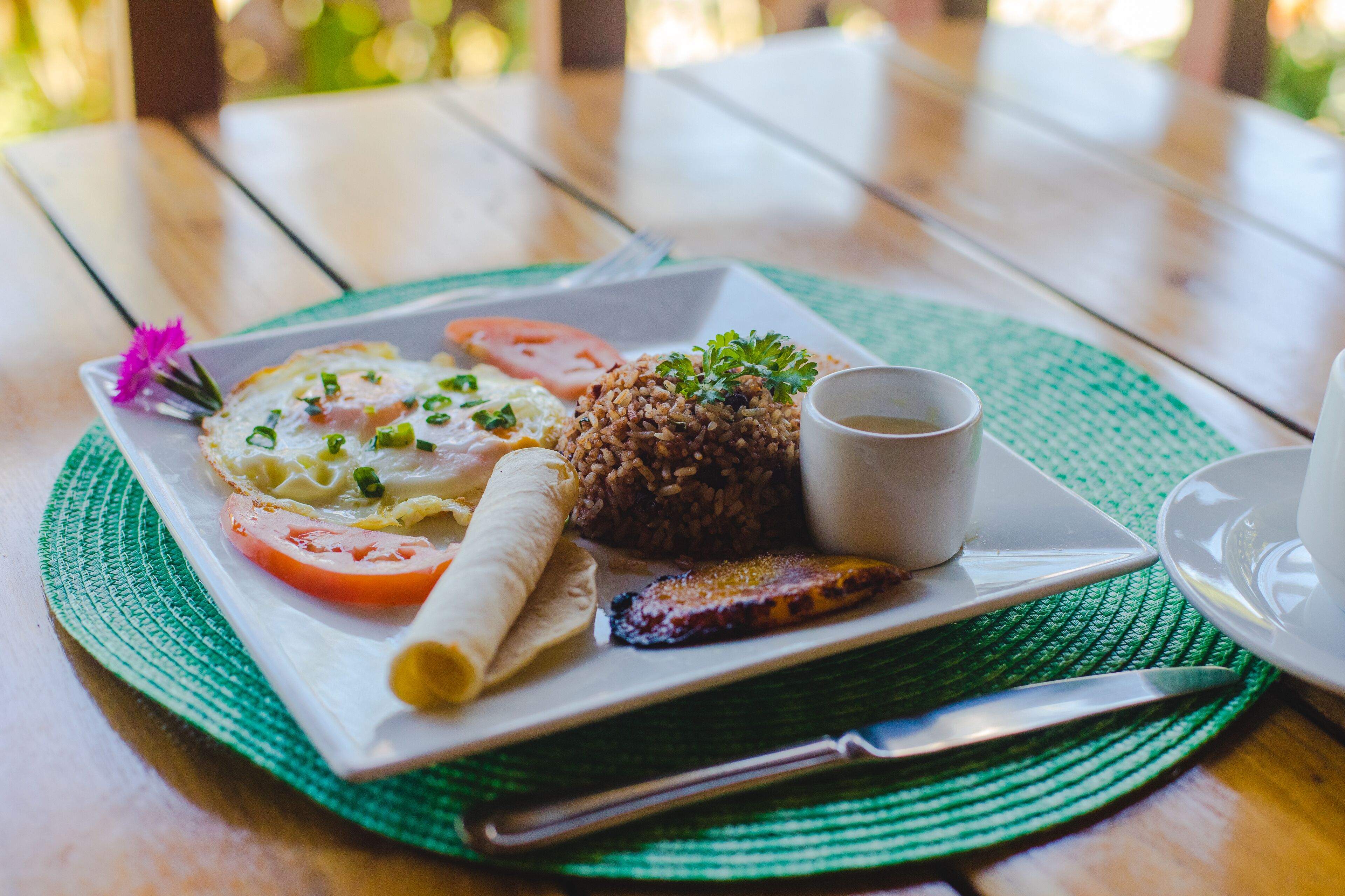 Uma refeição sobre mesa de madeira, com um jogo americano verde, incluindo arroz, um omelete enrolado, uma fatia de tomate e uma xícara de café.