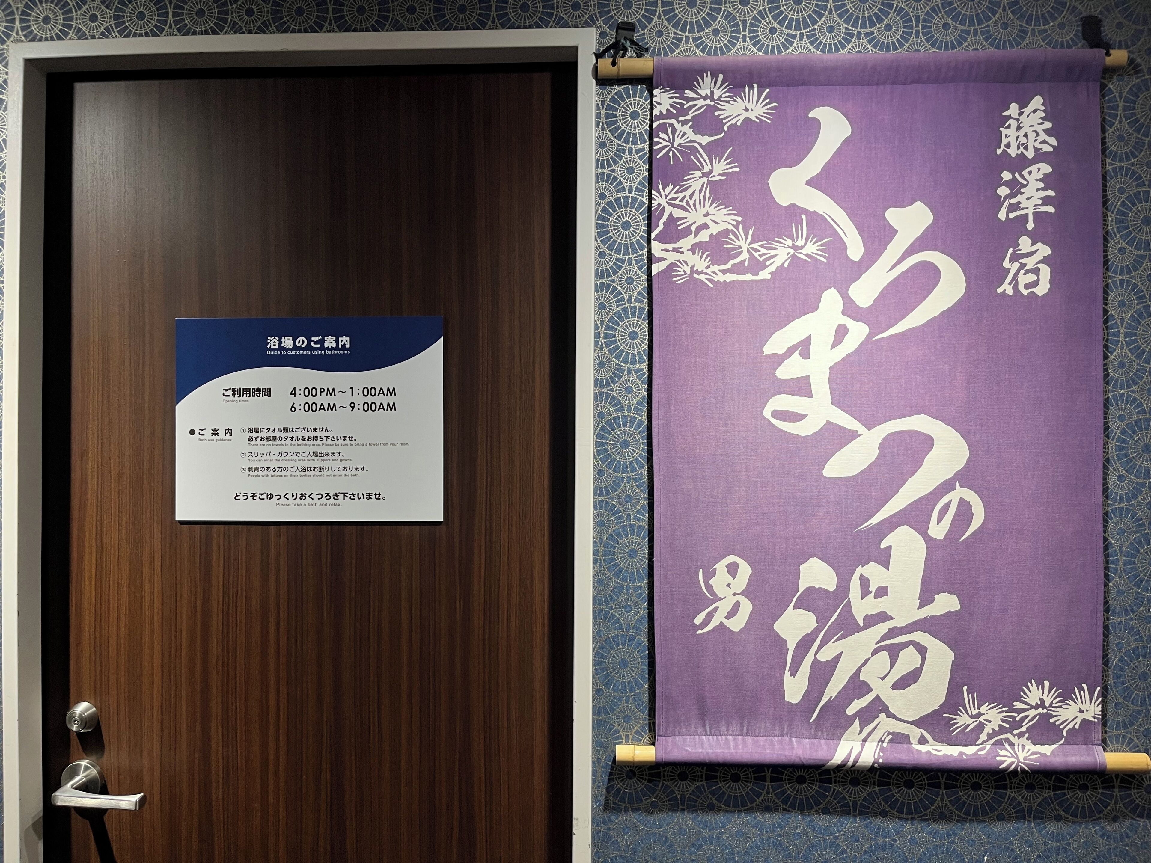 Quarto de hotel com porta de madeira, placa com horário de funcionamento e faixa roxa com caracteres japoneses.