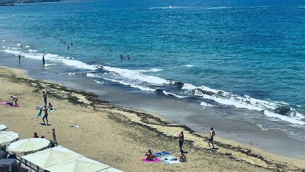 Uma praia com pessoas tomando sol e nadando, barracas montadas e uma vista clara do mar e colinas distantes.