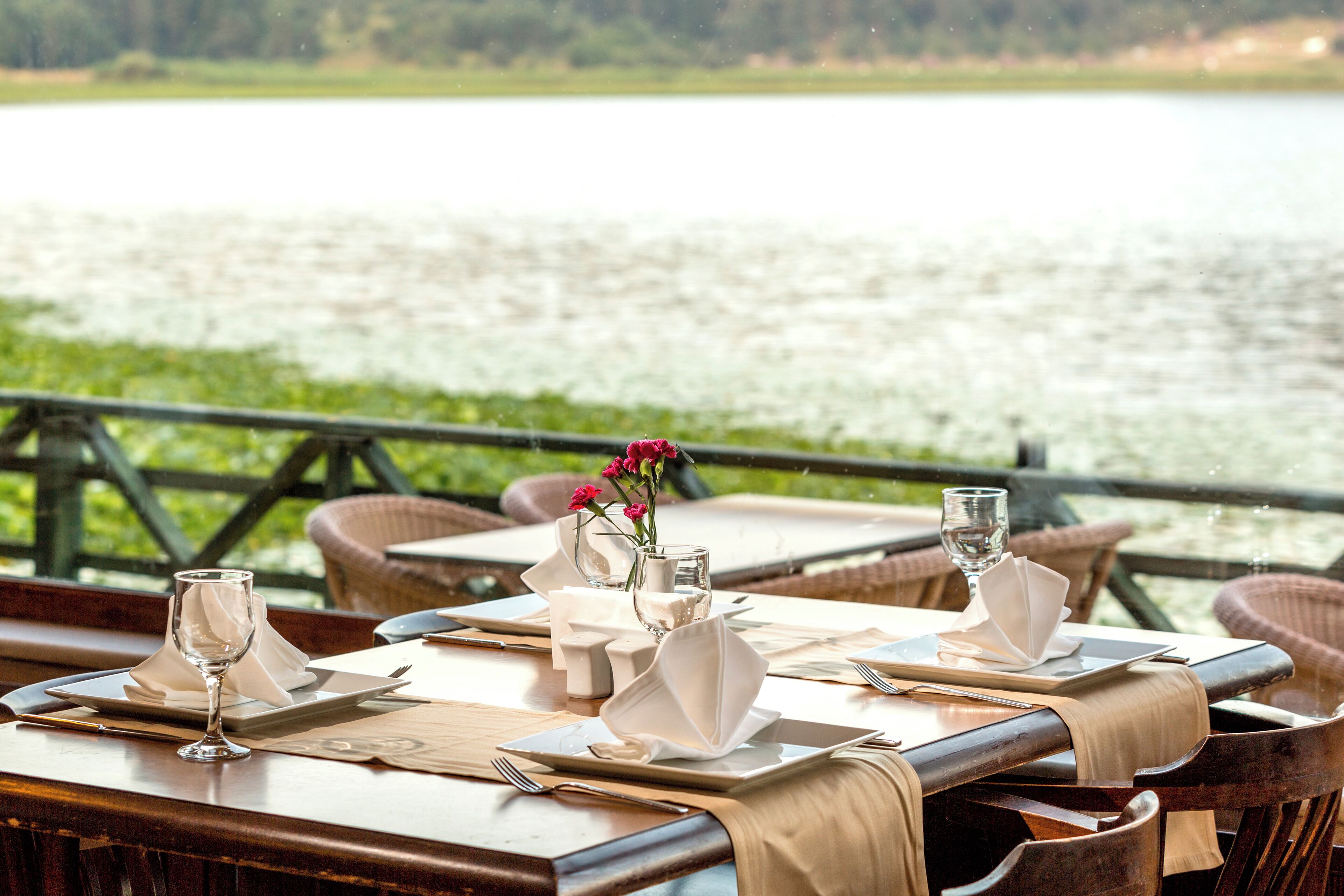 Mesa de jantar posta com guardanapos brancos, pratos e talheres, com vista para um lago.