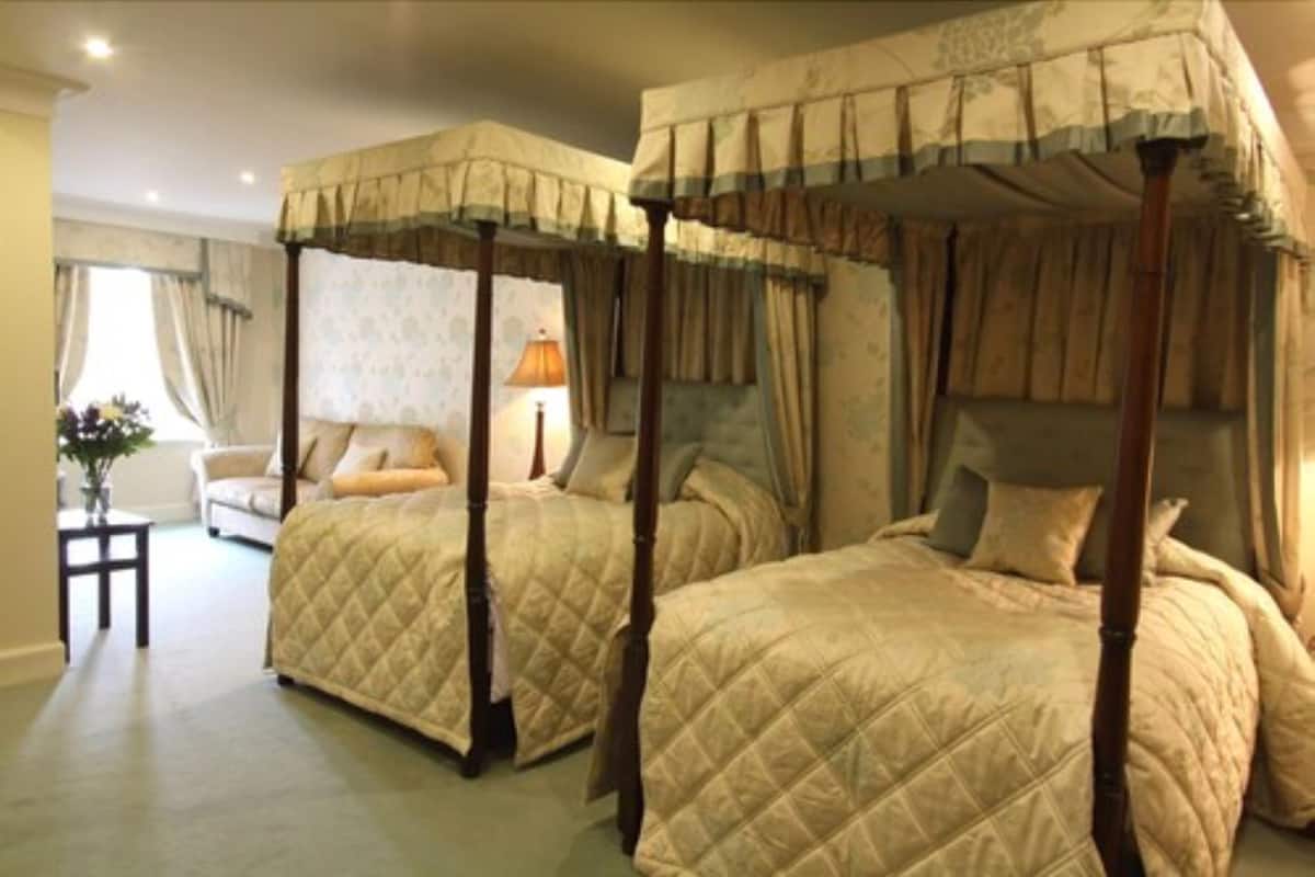 Uma cama com dossel, com colcha acolchoada, uma mesa de cabeceira com abajur e uma cadeira num quarto com carpete e cortinas.