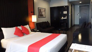 Quarto de hotel com cama, escrivaninha, cadeira, mesa de cabeceira e televisão na parede.