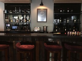 Uma área de bar com várias prateleiras de garrafas de bebidas, uma máquina de café e banquetas estofadas vermelhas.