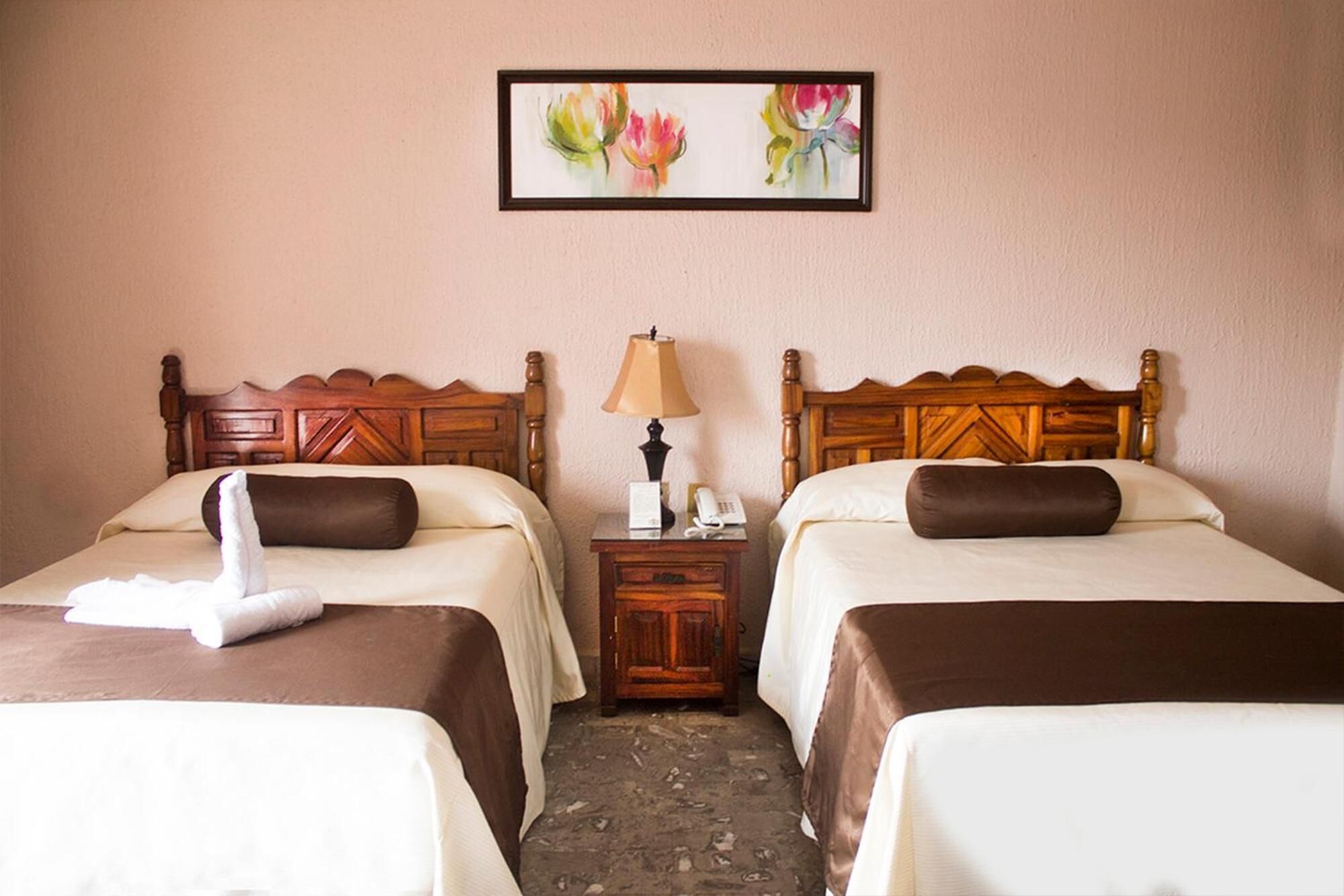 Quarto de hotel com duas camas, uma mesa de cabeceira com abajur e uma pintura floral emoldurada na parede.