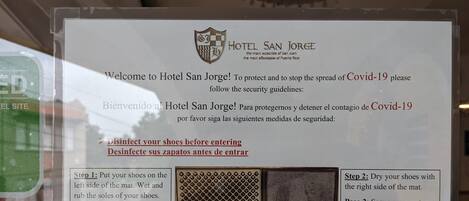 Placa no Hotel San Jorge detalhando medidas de segurança contra COVID-19 para hóspedes, incluindo desinfecção de sapatos.
