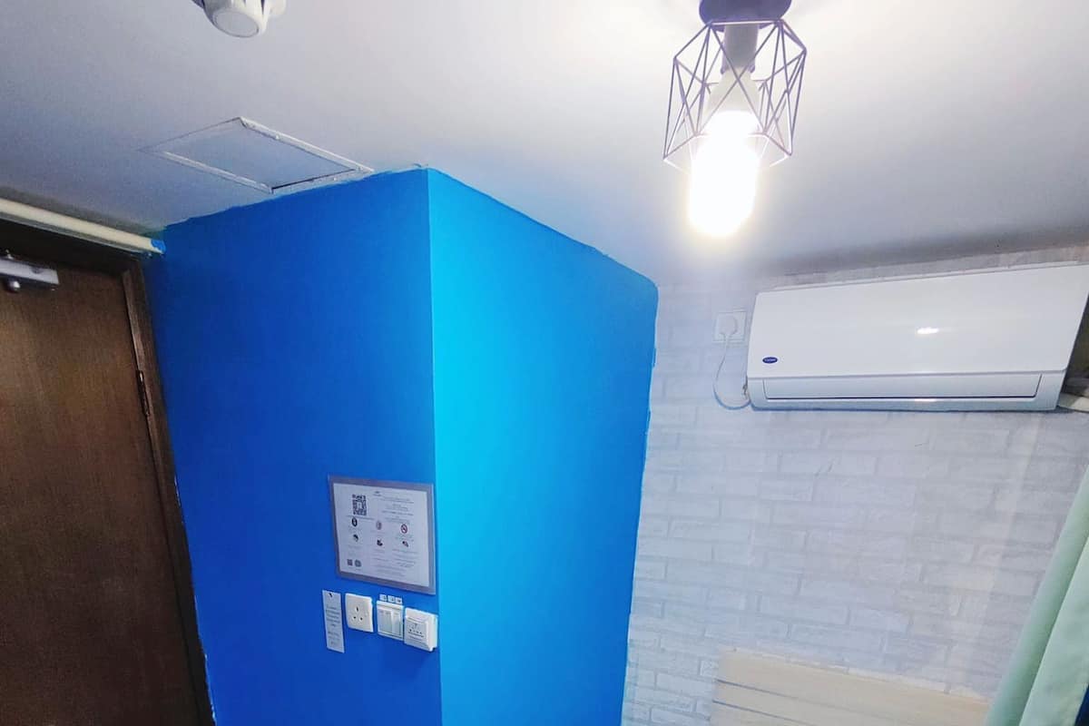 Quarto de hotel com uma parede azul de destaque, uma cama com roupa de cama branca, uma cabeceira de madeira, uma mesa de cabeceira, um ar condicionado fixado na parede e uma tomada.