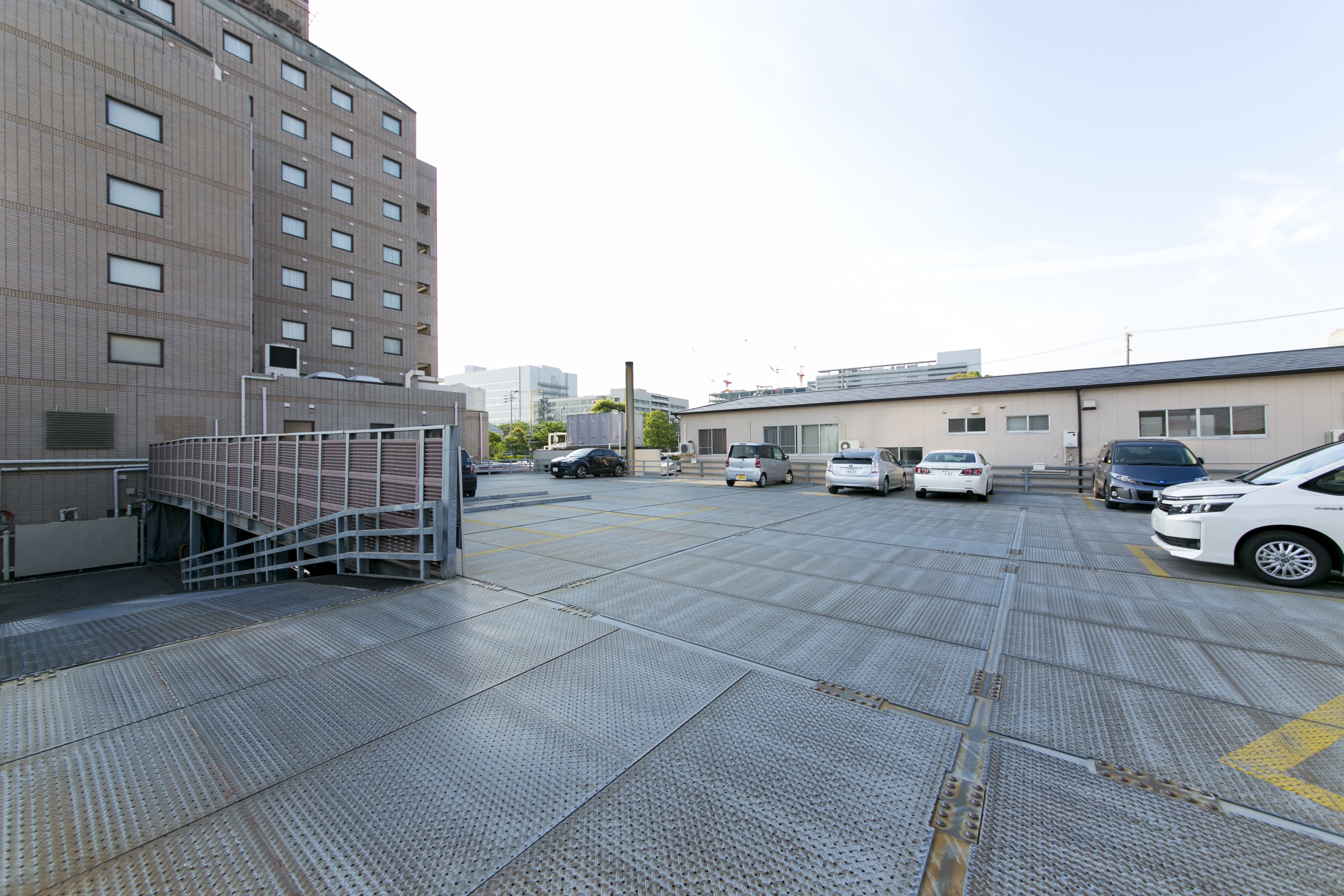 Um estacionamento com vários carros, um edifício com diversas janelas e uma escadaria que leva a uma entrada.