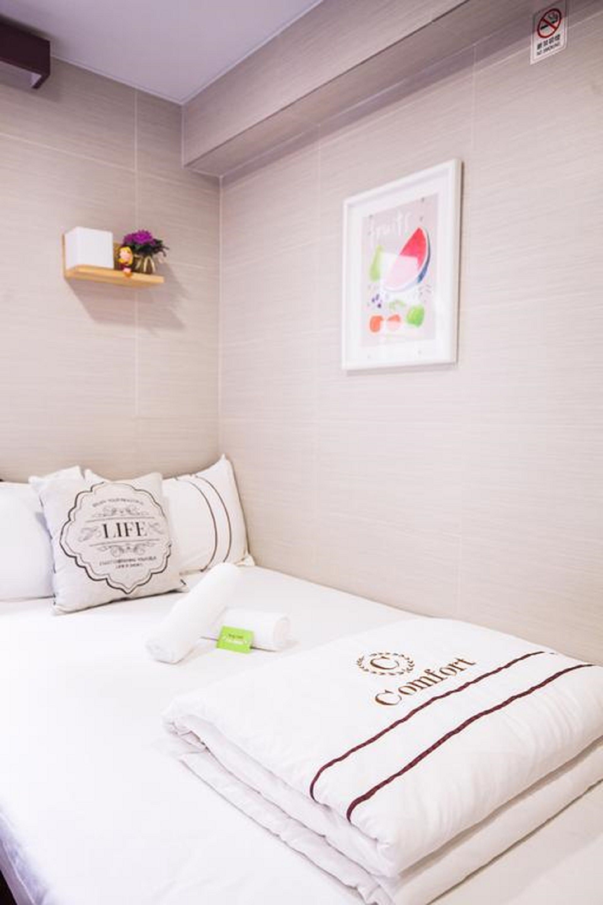 Cama bem arrumada com edredom branco e travesseiros, uma pequena estante com um vaso de flores e um quadro na parede.