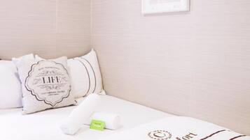 Cama bem arrumada com edredom branco e travesseiros, uma pequena estante com um vaso de flores e um quadro na parede.