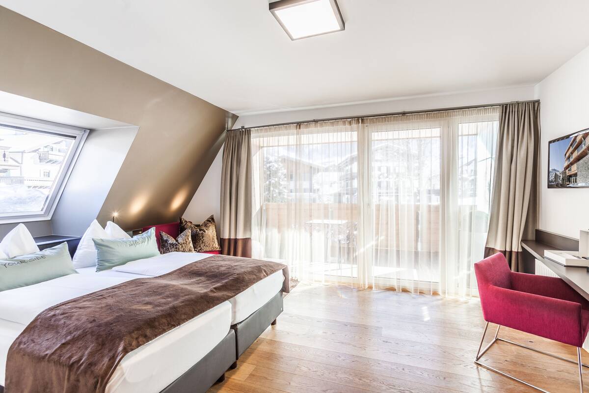 Quarto de hotel moderno com uma cama grande, uma mesa de trabalho e uma cadeira. Há uma janela com cortinas, uma luminária no teto e uma pintura na parede.
