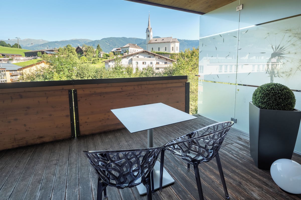 Uma varanda com mesa e cadeiras, um vaso de planta e vista para uma igreja e montanhas.