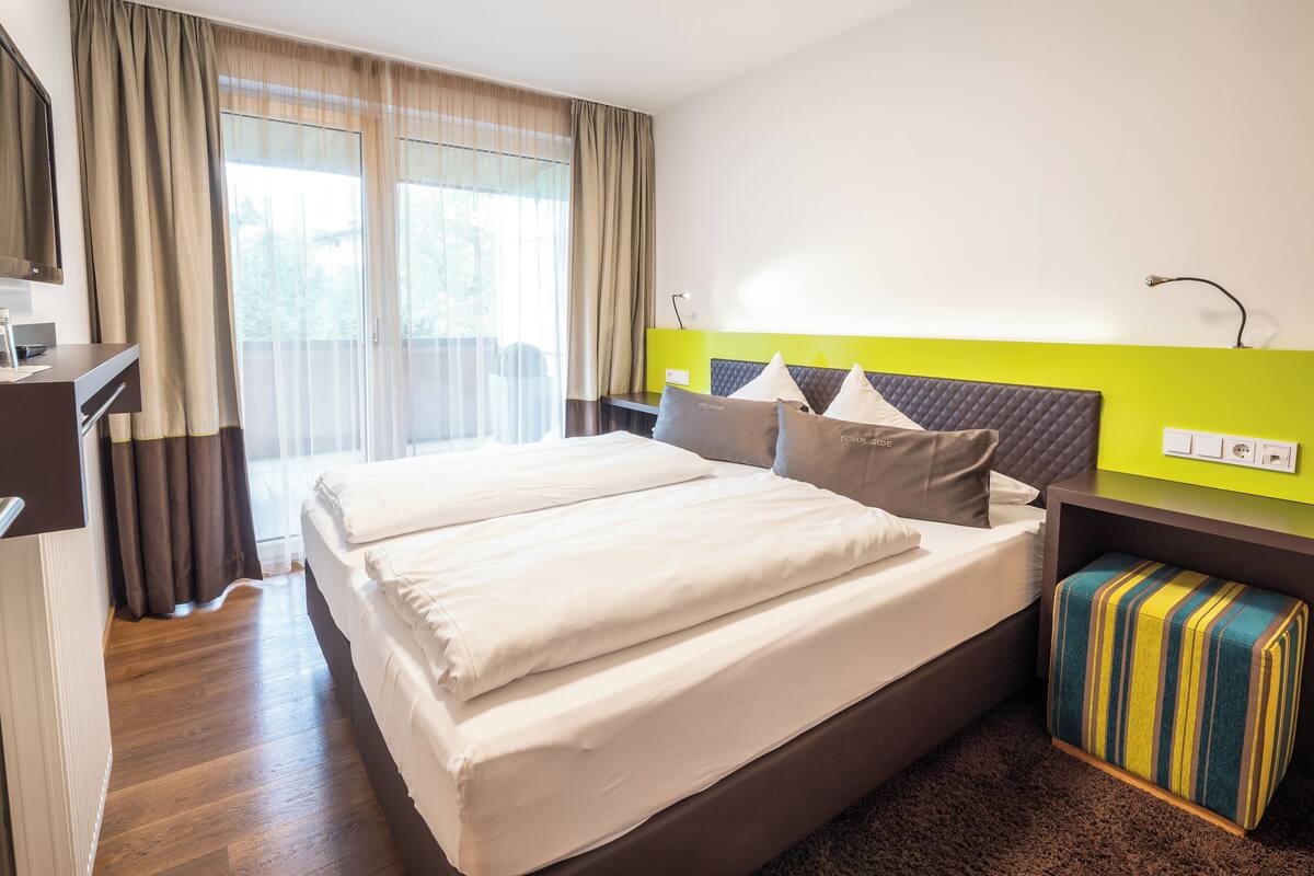 Quarto de hotel com uma cama grande, mesa de cabeceira, uma cadeira e uma janela com cortinas.